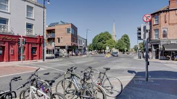 Vista da rua com bicicletas estacionadas na calçada, prédios com lojas e o topo de uma igreja ao fundo.