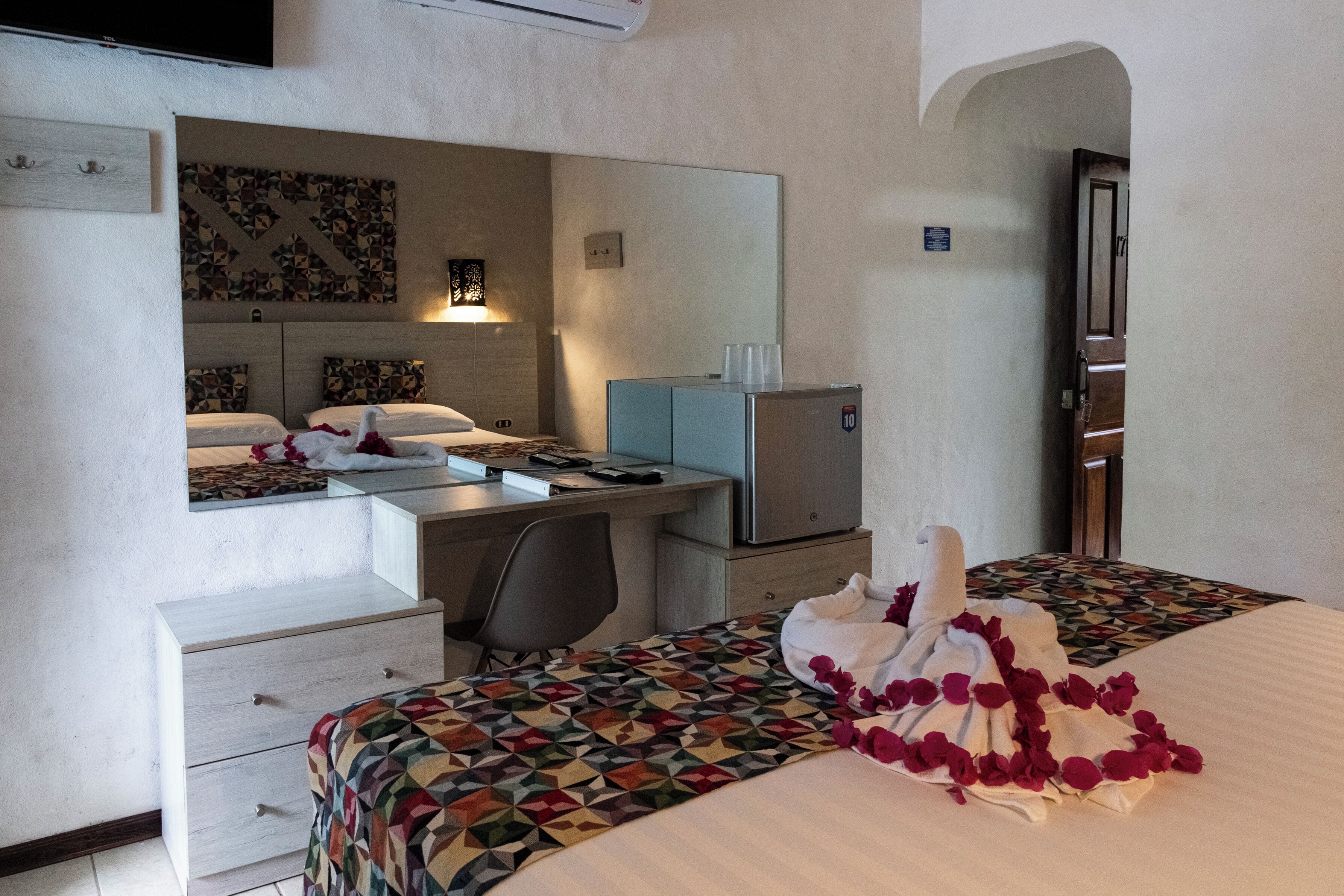 Quarto de hotel com uma cama, uma escrivaninha, uma cadeira, um pequeno frigorífico, um espelho e uma porta.