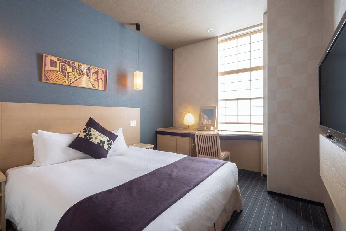 Quarto de hotel com uma cama grande, uma escrivaninha, uma cadeira, uma janela e obras de arte na parede.
