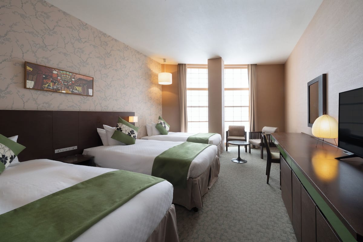 Quarto de hotel com duas camas, uma escrivaninha com televisão, uma cadeira e uma janela com cortinas.
