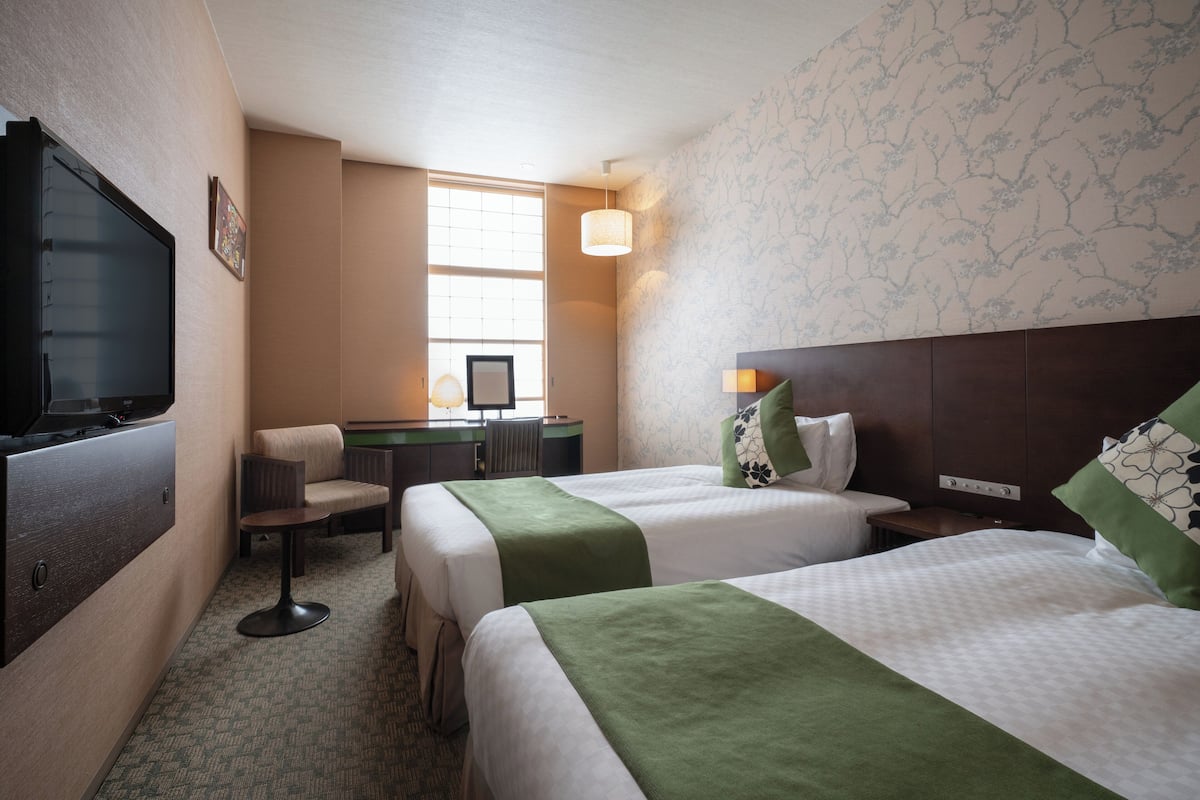Quarto de hotel com duas camas, uma escrivaninha, uma cadeira, uma televisão e uma janela.