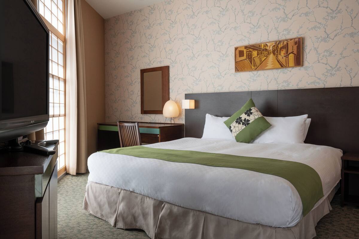 Quarto de hotel com uma cama grande, uma escrivaninha, uma televisão e uma janela com cortinas.