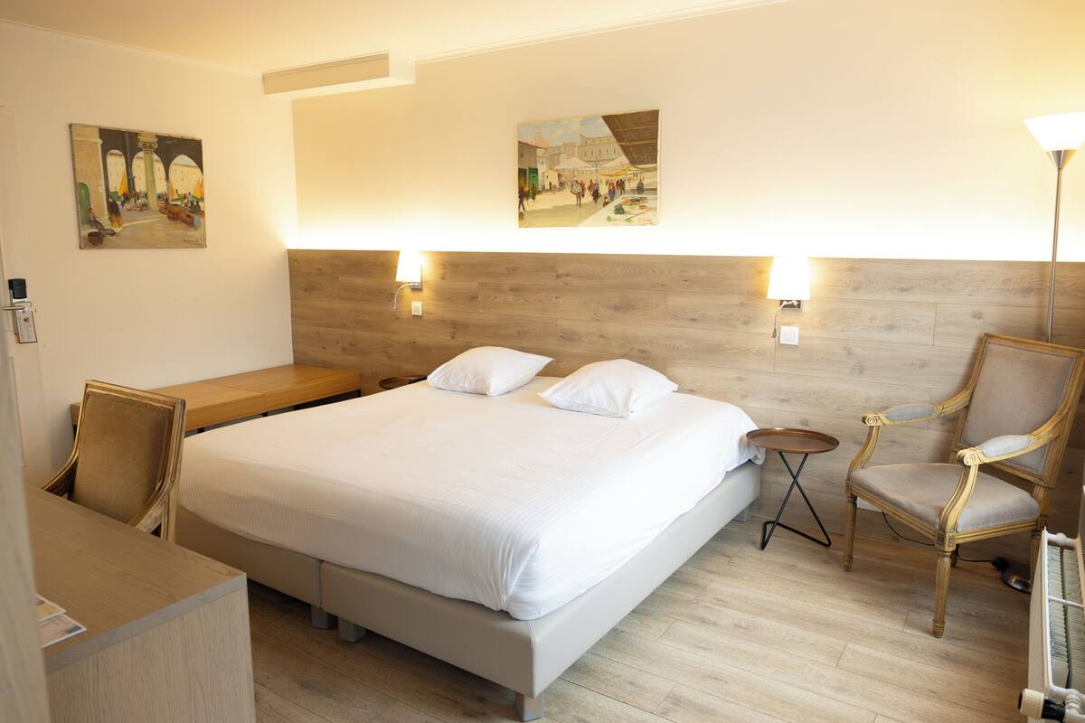 Quarto de hotel com uma cama grande, uma escrivaninha, uma cadeira e duas pinturas na parede.