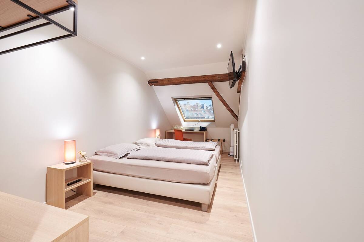 Quarto moderno com teto inclinado, cama com duas almofadas, mesa de cabeceira com abajur e escrivaninha com computador.