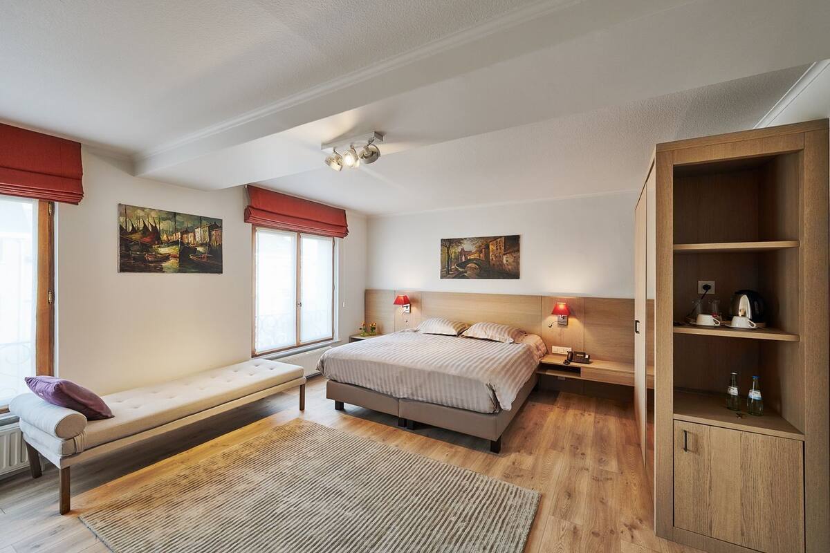 Quarto com cama, mesas de cabeceira, um banquinho, armário de madeira e obras de arte nas paredes.
