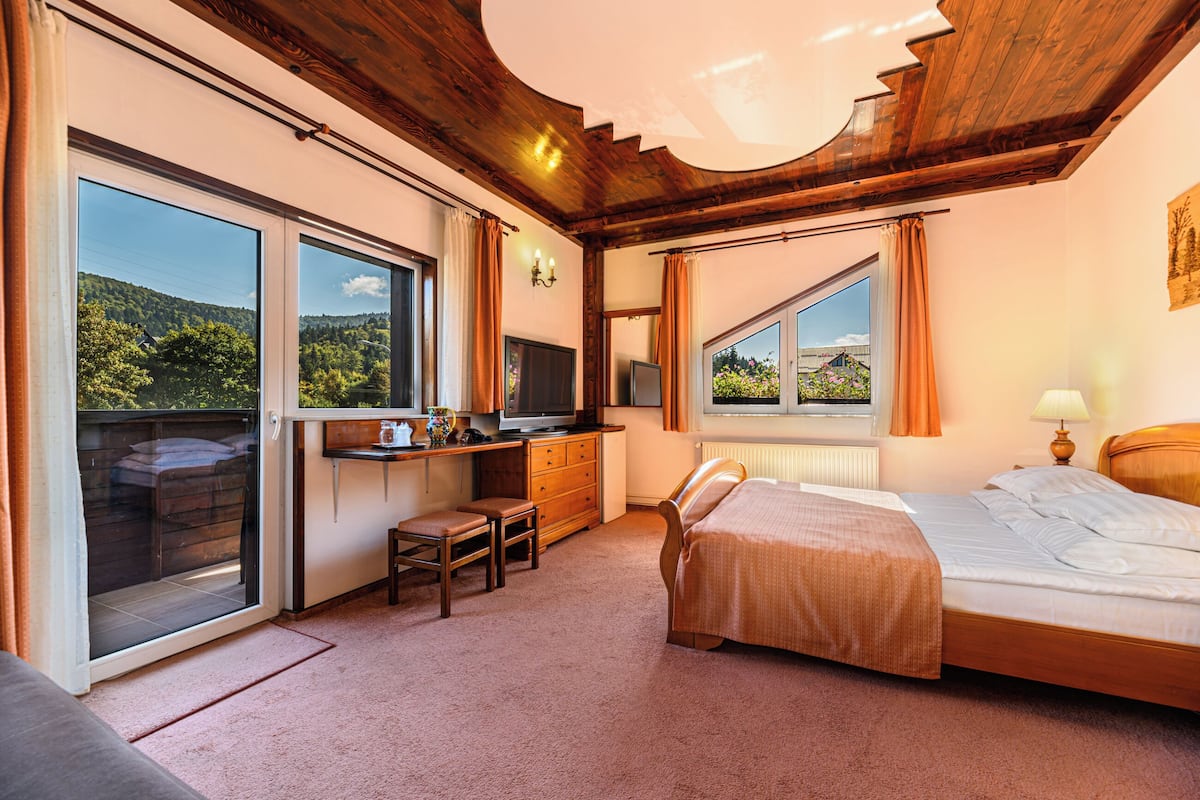 Quarto com cama, escrivaninha, cadeira, televisão e janelas amplas.
