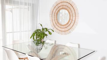 Uma área de refeições moderna com mesa de vidro, cadeiras brancas, ventilador de teto e um elemento decorativo na parede.