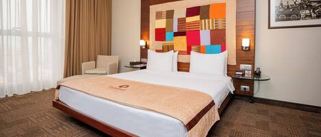 Quarto de hotel com uma cama grande, cabeceira de madeira, colcha colorida, mesa de cabeceira, luminária e um quadro na parede.