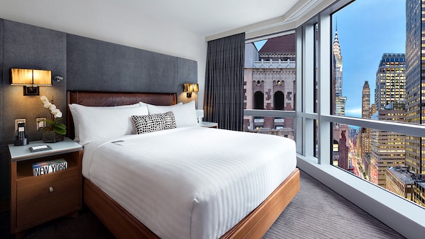 Quarto de hotel com cama grande, mesa de cabeceira, luminária e vista para a cidade.