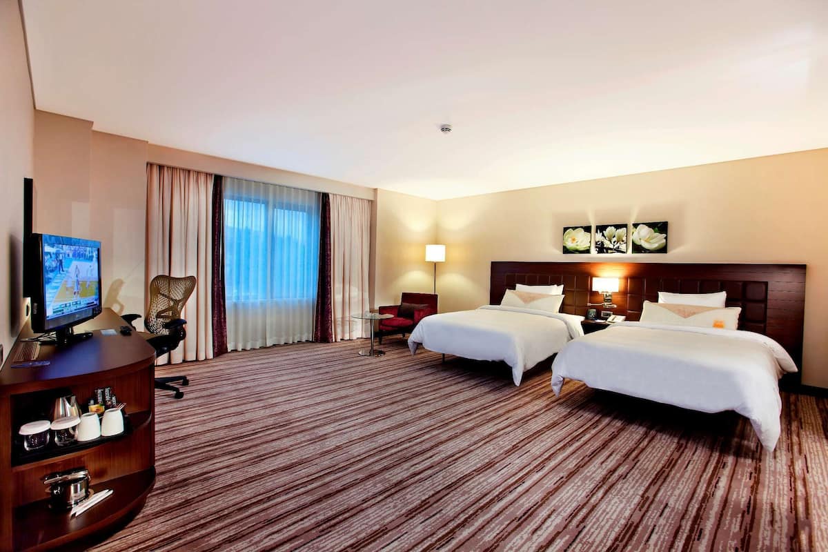 Quarto de hotel com duas camas, televisão, escrivaninha com abajur e uma janela grande com cortinas.