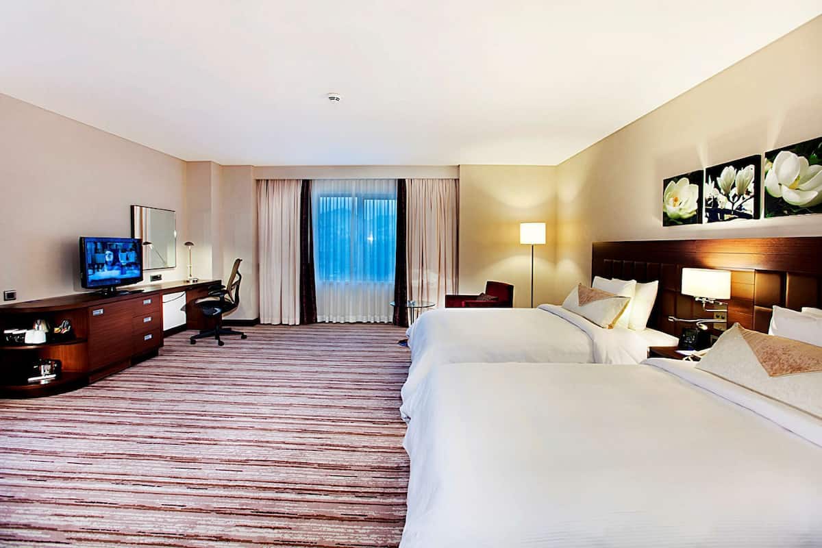 Quarto de hotel com duas camas, uma escrivaninha com televisão, uma cadeira e uma janela grande com cortinas.