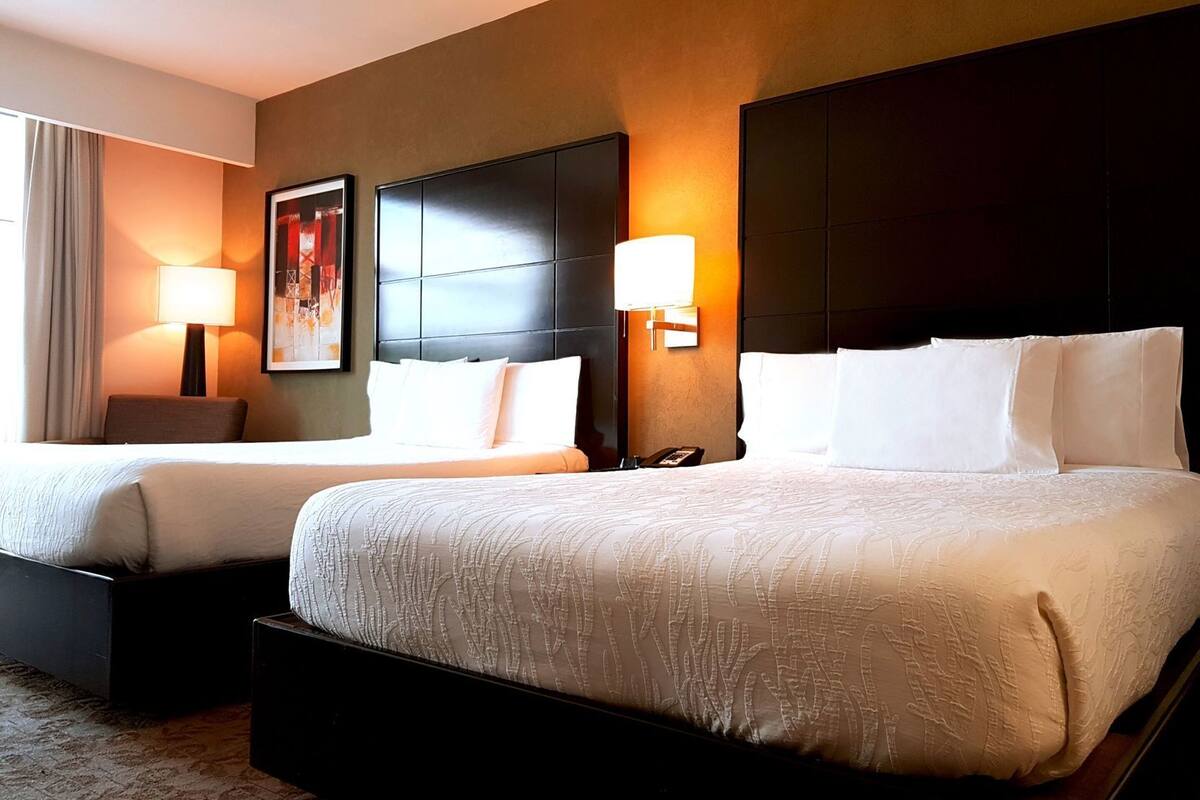 Quarto de hotel com duas camas, cabeceira com luminária, um quadro na parede e um abajur próximo à janela.