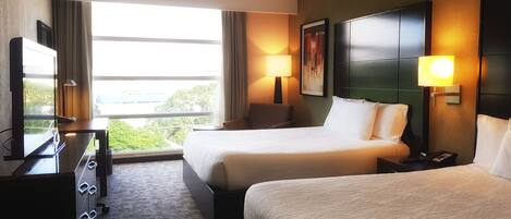 Quarto de hotel com duas camas, televisão, escrivaninha e janela com vista para vegetação.