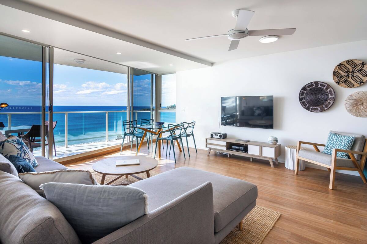 Uma sala de estar moderna com uma TV grande, assentos confortáveis e uma varanda com vista para o oceano.