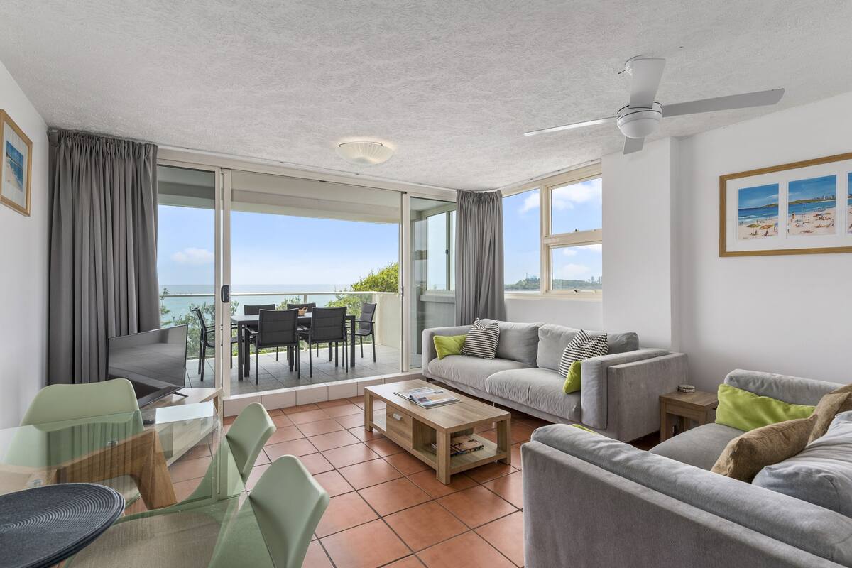 Uma sala de estar moderna com sofá, mesa de centro e cadeiras. Há uma varanda com mesa e cadeiras, e vista para o mar.