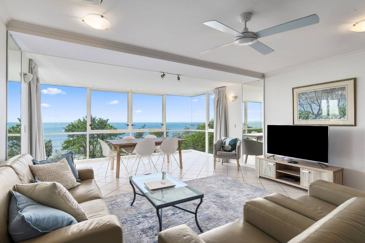 Uma sala de estar moderna com uma televisão grande, assentos confortáveis, um espaço de refeições e uma varanda com vista para o mar.