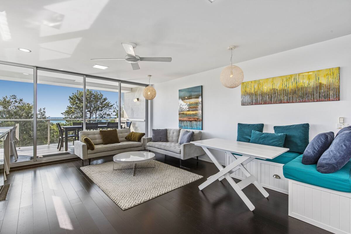 Uma sala de estar moderna com sofá, mesa de centro e área de jantar. Há uma grande janela com vista para o exterior.
