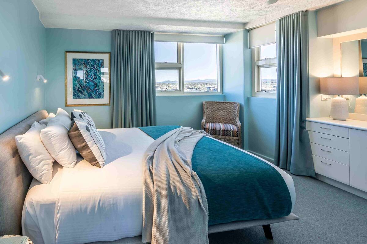Quarto de hotel com cama, poltrona, escrivaninha e janela com cortinas.