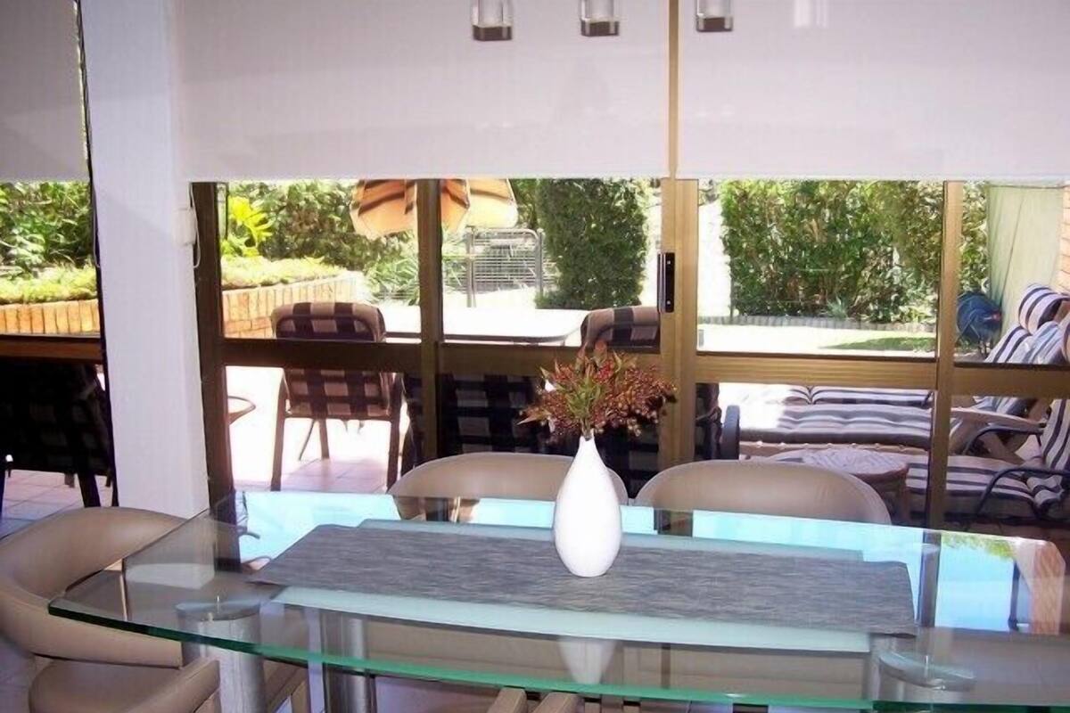 Uma sala de jantar moderna com mesa e cadeiras de vidro, um vaso com flores e vista para o pátio externo.