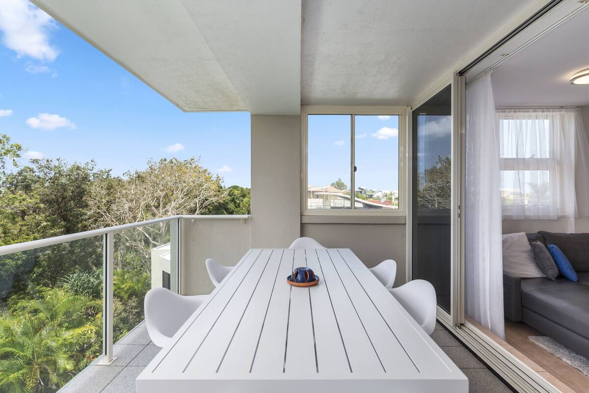 Uma varanda com mesa e cadeiras brancas, vista para o verde e uma porta de vidro deslizante que dá para uma sala de estar com sofá.