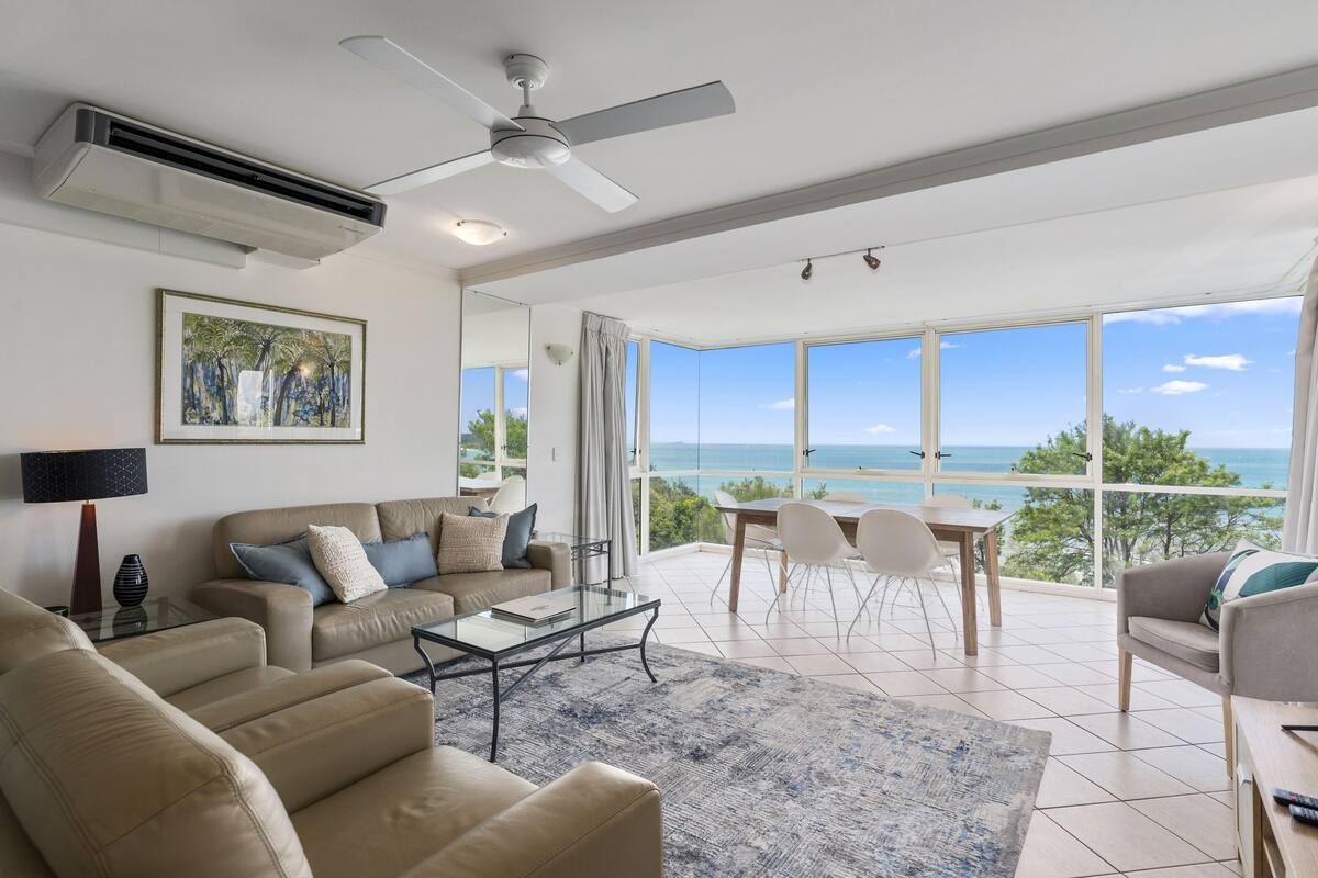 Uma sala de estar ampla com uma grande janela que oferece vista para o mar, um ventilador de teto e um espaço de jantar com mesa e cadeiras.