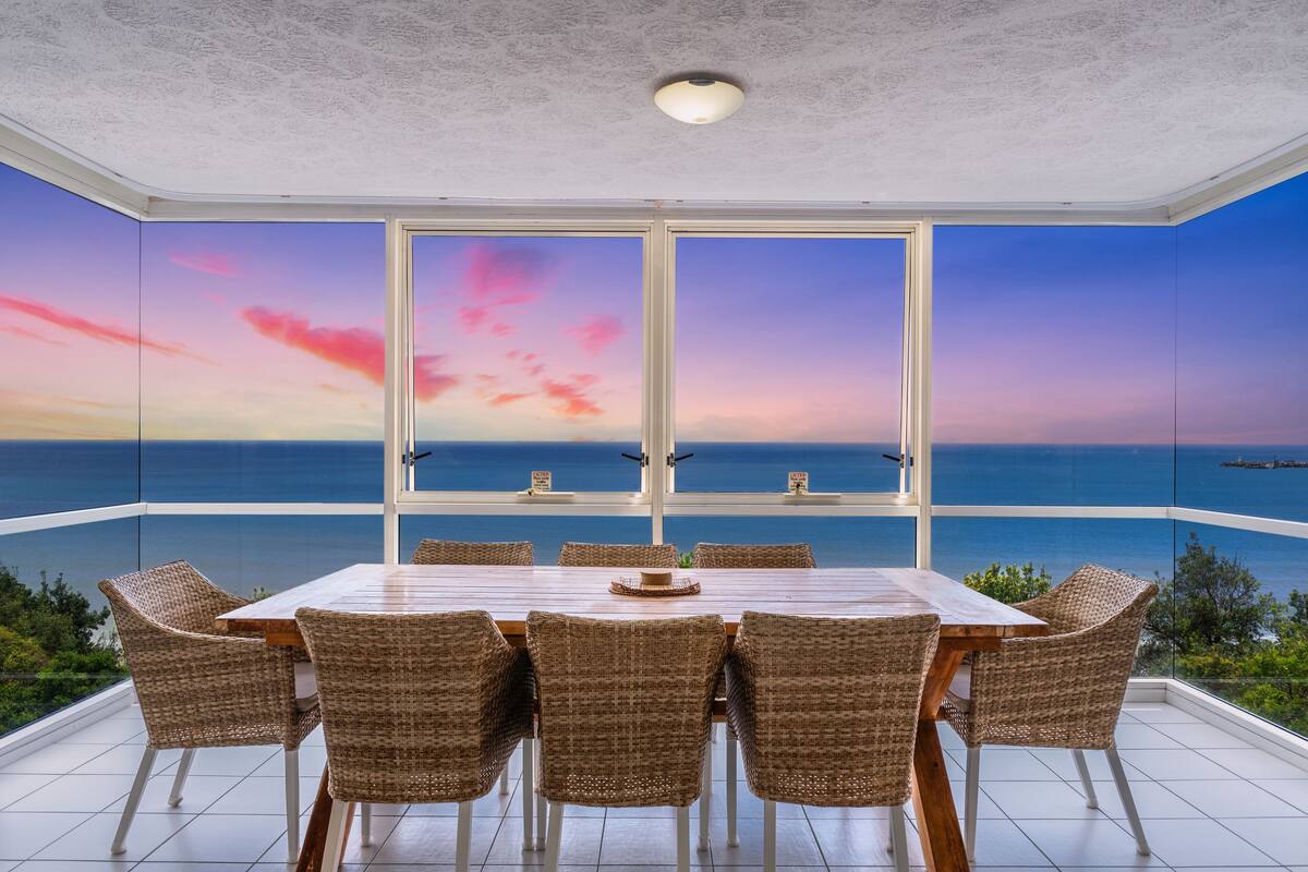 Uma área de refeições com mesa e cadeiras, janelas amplas com vista para o mar e uma luminária no teto.
