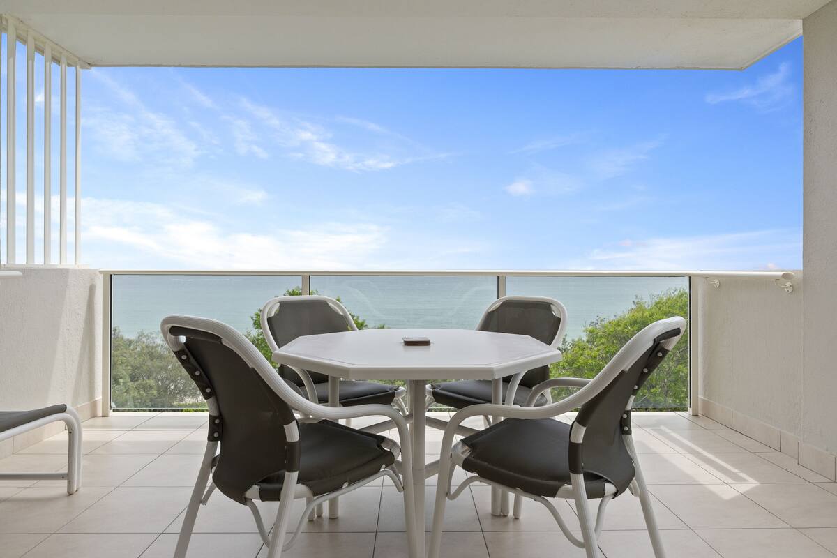 Varanda com mesa branca e quatro cadeiras, com vista panorâmica para o mar e vegetação.