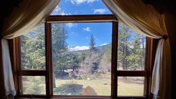 Janela com cortinas, com vista para uma paisagem arborizada e montanhas ao fundo.