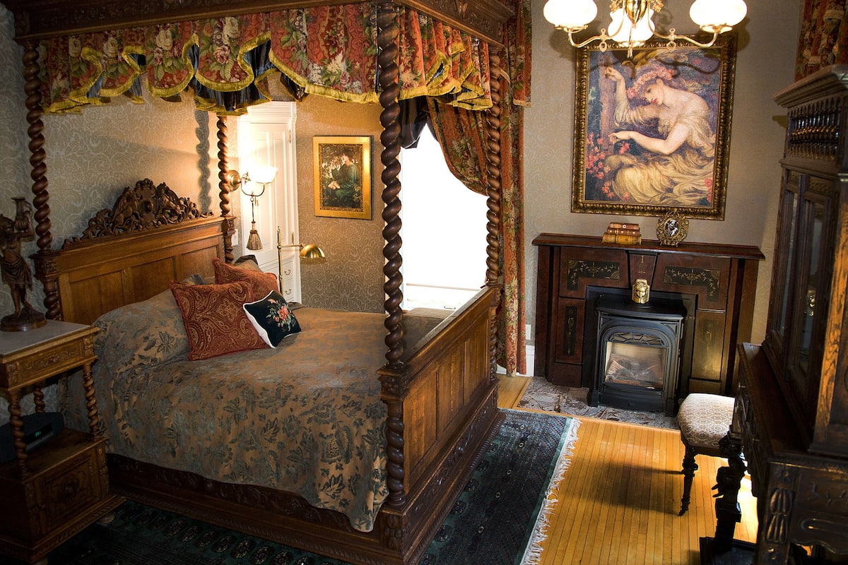 Uma cama com dossel e cortina floral, lareira e um quadro na parede.
