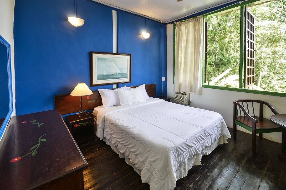 Quarto de hotel com cama, mesa de cabeceira, luminária, cadeira e janela com vista para áreas verdes.
