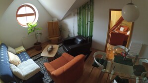 Uma sala de estar estilo loft com teto inclinado, uma janela redonda, um sofá, uma mesa de centro, uma cadeira, uma mesa de jantar de vidro e uma planta de bambu.