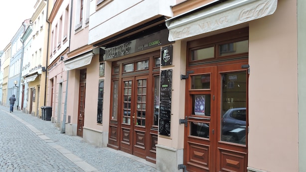 Uma rua de paralelepípedos ladeada por edifícios, incluindo um bar chamado 'New Age' com portas de madeira e um letreiro da Coca-Cola.