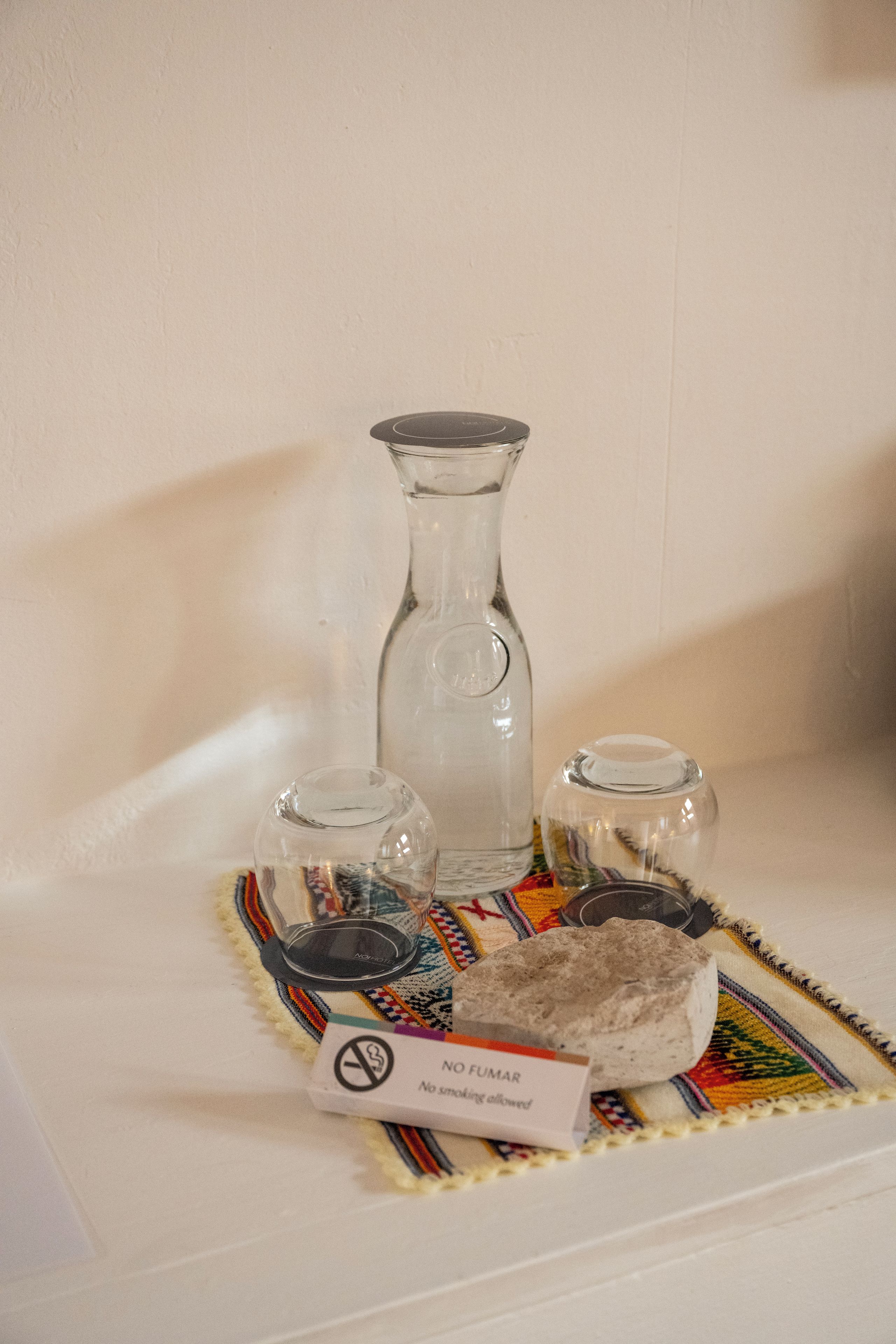 Uma garrafa de vidro transparente, dois recipientes de vidro menores e uma pedra com o aviso 'Não permitido fumar' sobre um tecido estampado.