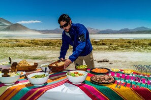 Alguém servindo comida sobre um cobertor colorido em uma paisagem aberta e extensa, com montanhas ao fundo.
