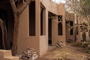 Um edifício com vários quartos, cada um com janela e porta. Árvores e pedras em frente ao edifício.