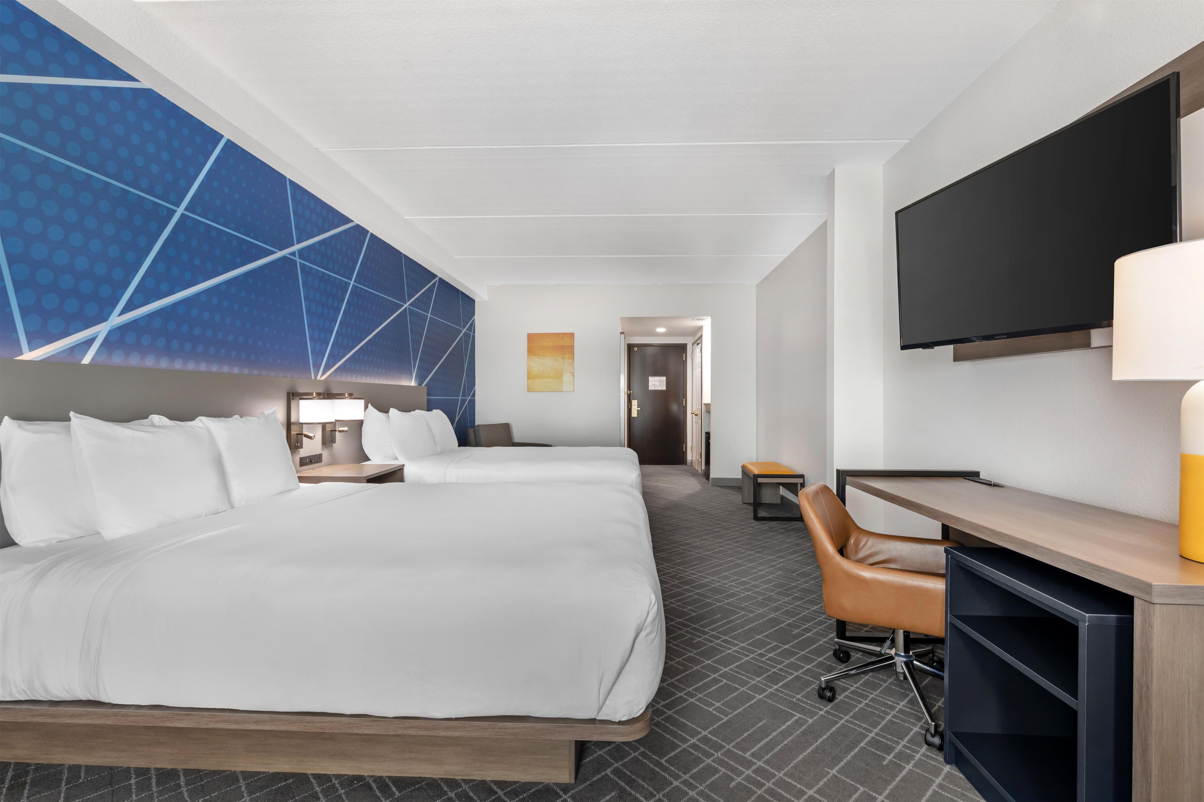 Quarto de hotel com duas camas, uma escrivaninha, uma cadeira, uma televisão e uma grande obra de arte na parede.