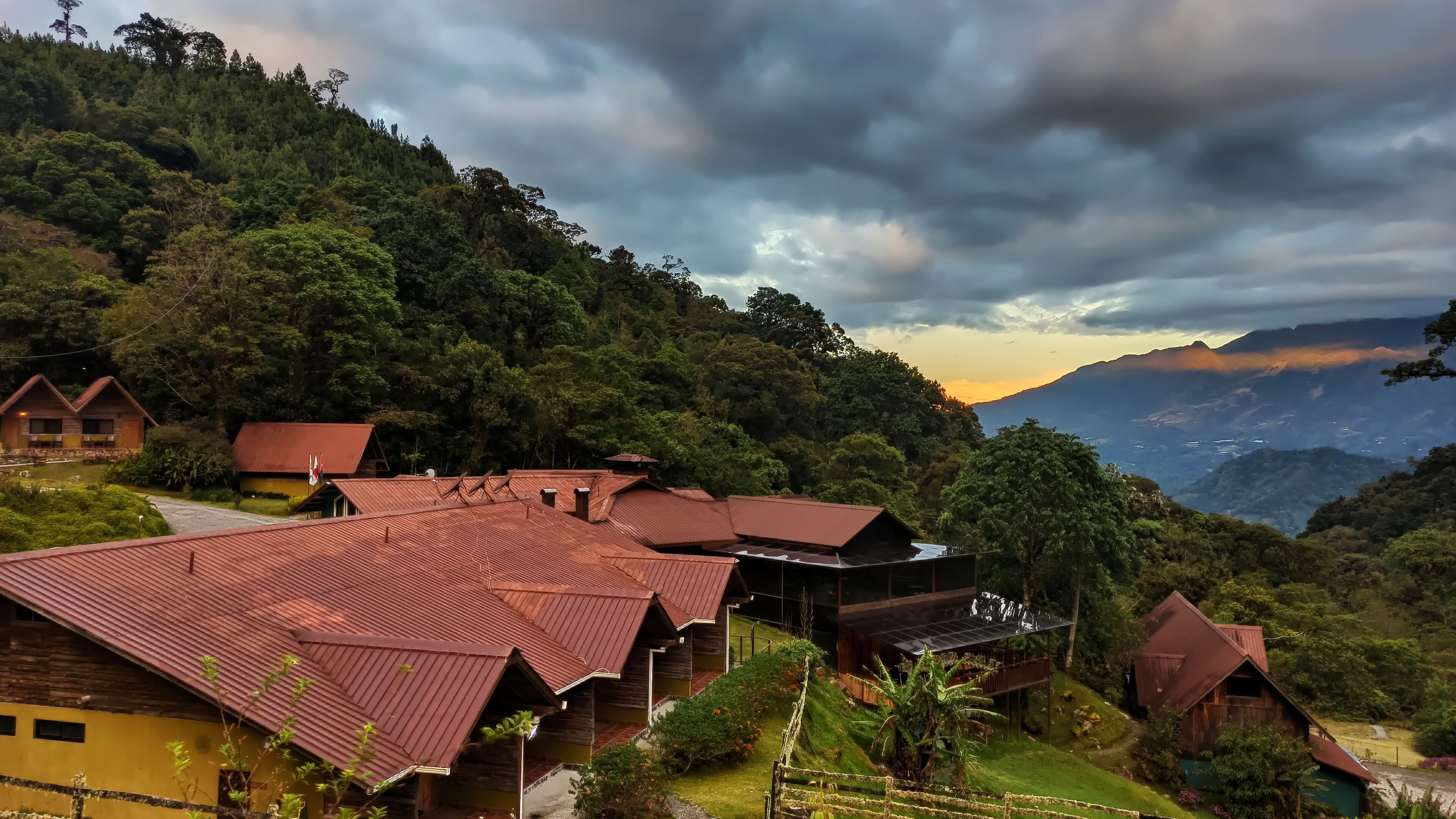 Um conjunto de cabanas rústicas com telhados vermelhos, cercadas por vegetação exuberante e montanhas ao fundo.