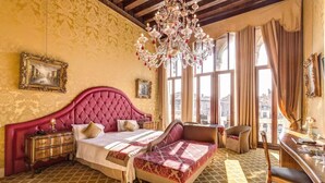 Quarto com uma cama grande, uma escrivaninha e uma cadeira. Há duas janelas com cortinas, um candelabro e um chão estampado.