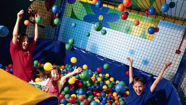 Espaço de recreação com tela de proteção, bolas coloridas e piso acolchoado azul e amarelo.