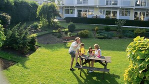 Uma família jantando em uma mesa ao ar livre em um jardim bem cuidado, com um edifício de dois andares ao fundo.