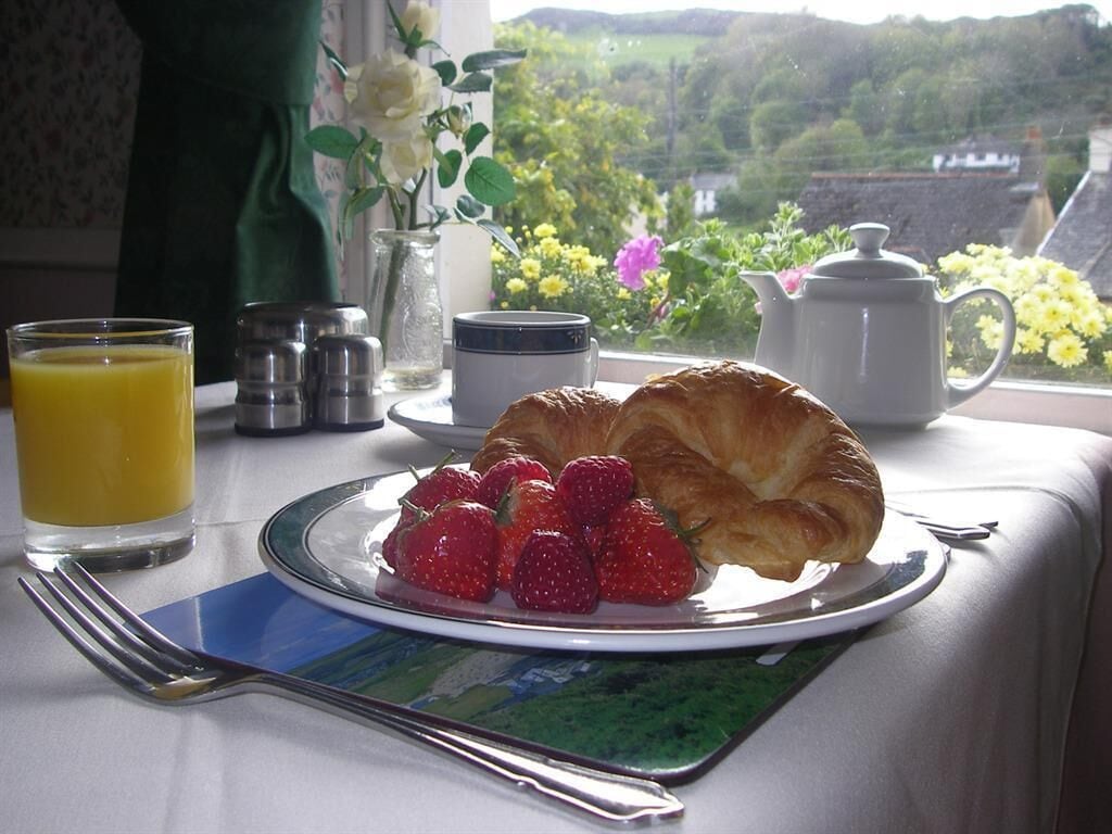 Disposição para o café da manhã com croissants, morangos e um copo de suco de laranja sobre uma mesa com vista para o verde e flores.