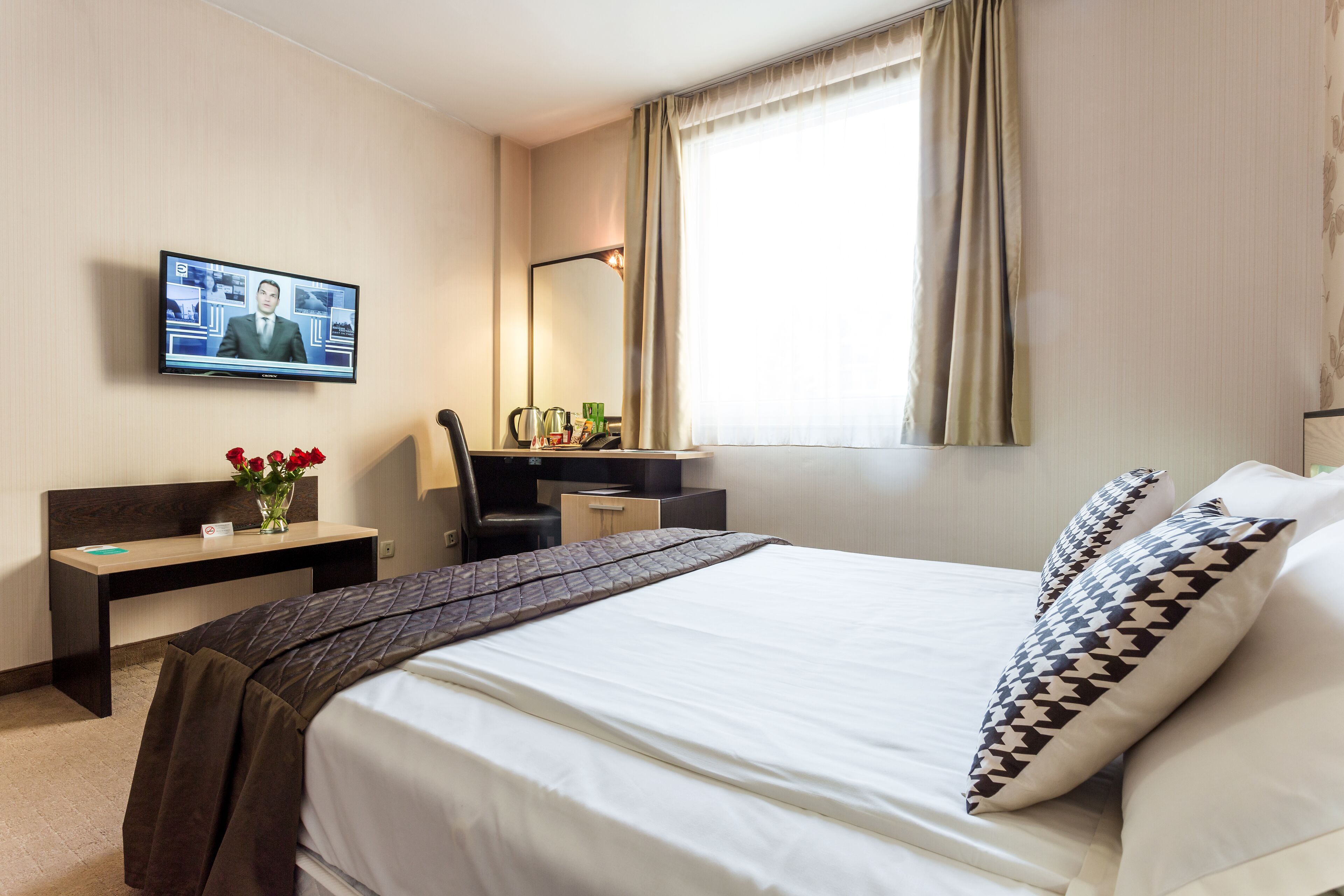 Quarto de hotel com uma cama, uma escrivaninha, uma cadeira, uma TV na parede e uma janela com cortinas.