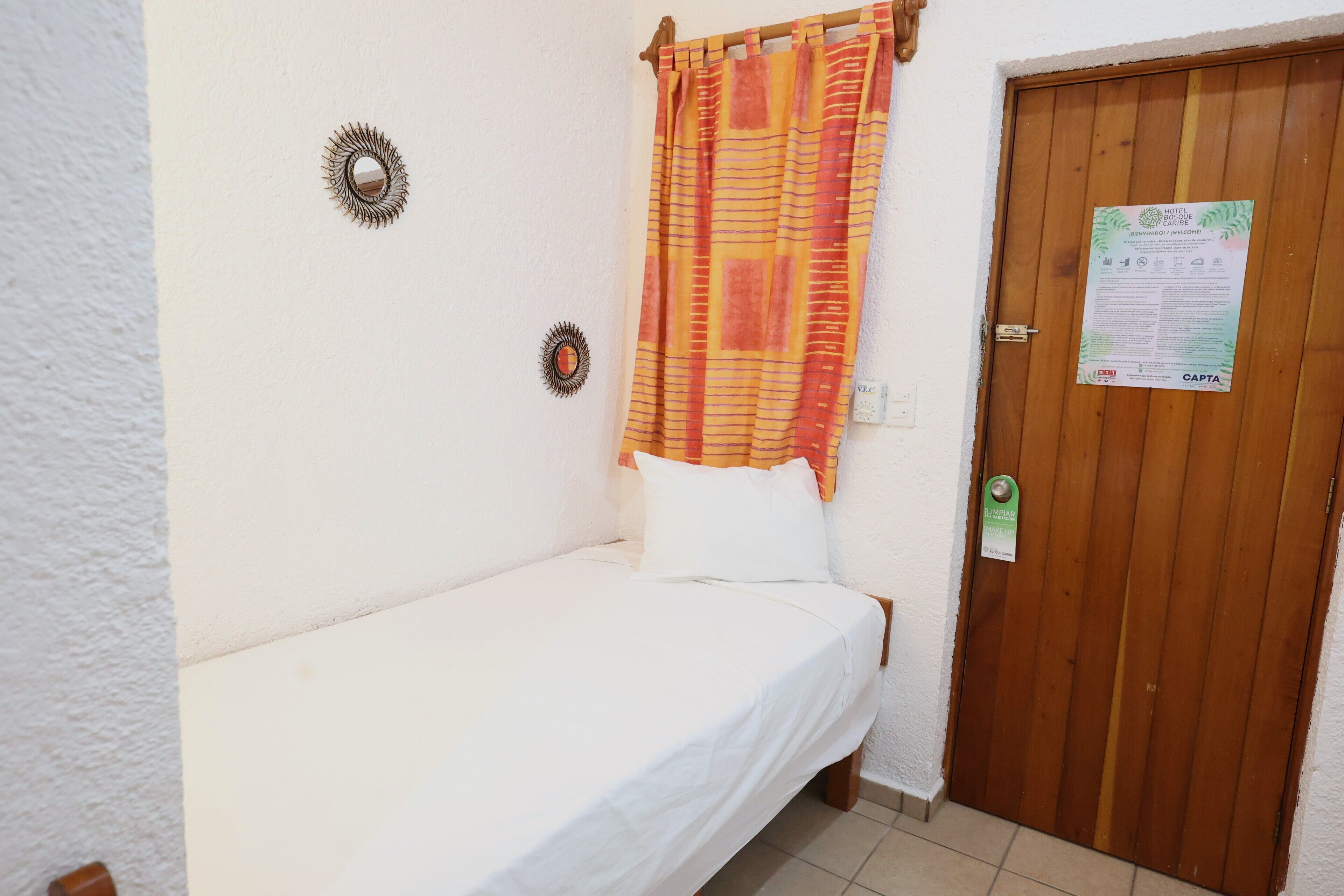 Uma cama de solteiro com roupa de cama branca, uma porta de madeira com um letreiro verde e dois espelhos decorativos fixados na parede.