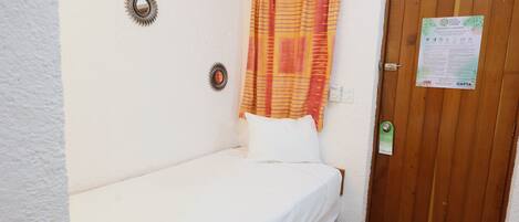 Uma cama de solteiro com roupa de cama branca, uma porta de madeira com um letreiro verde e dois espelhos decorativos fixados na parede.