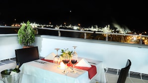 Mesa posta para dois com taças de vinho, uma garrafa e um pequeno vaso de planta numa varanda com vista para a cidade à noite.