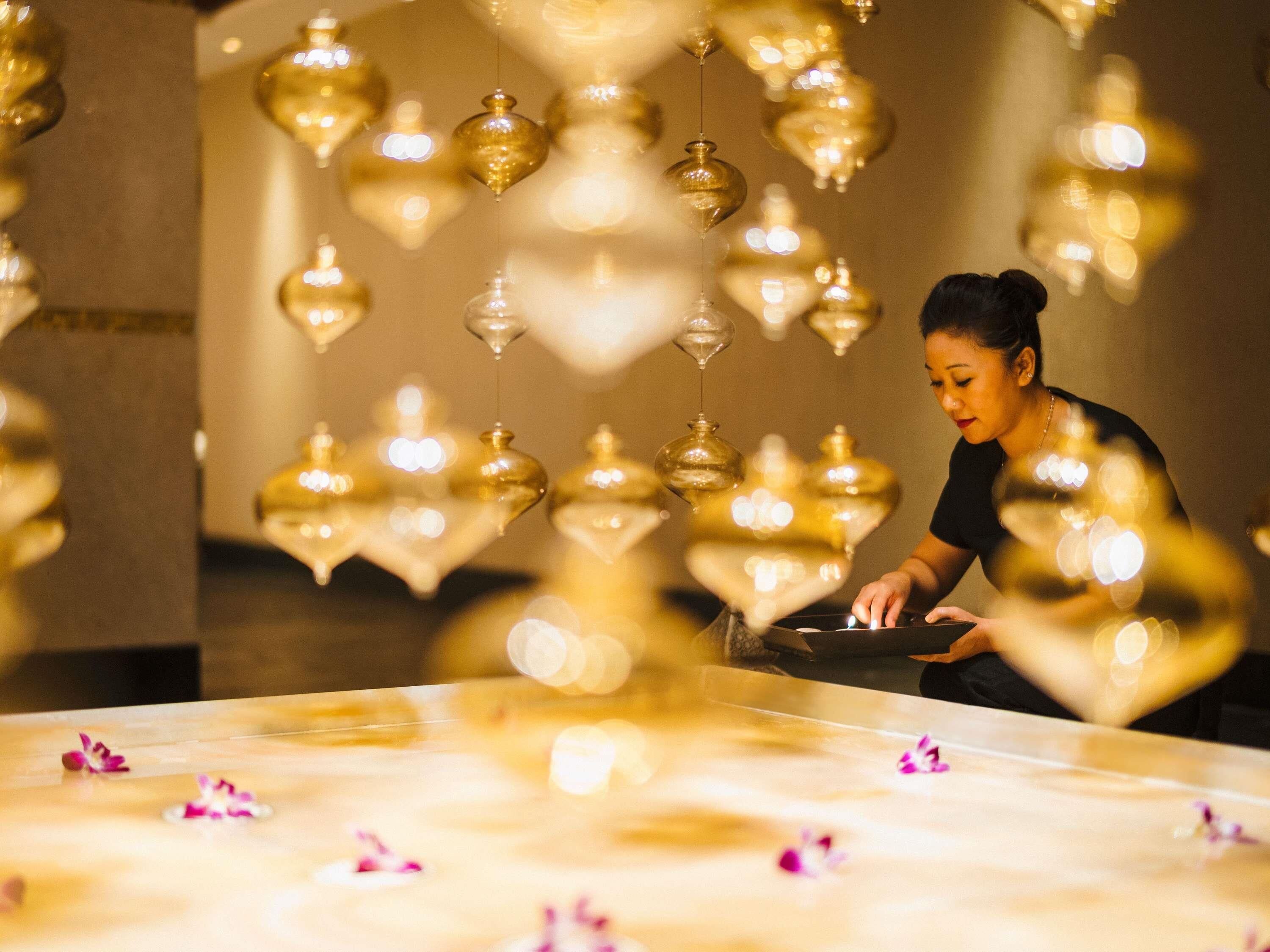 Uma pessoa trabalhando em uma mesa com um laptop, rodeada por luminárias pendentes e uma mesa com pétalas de flores espalhadas.