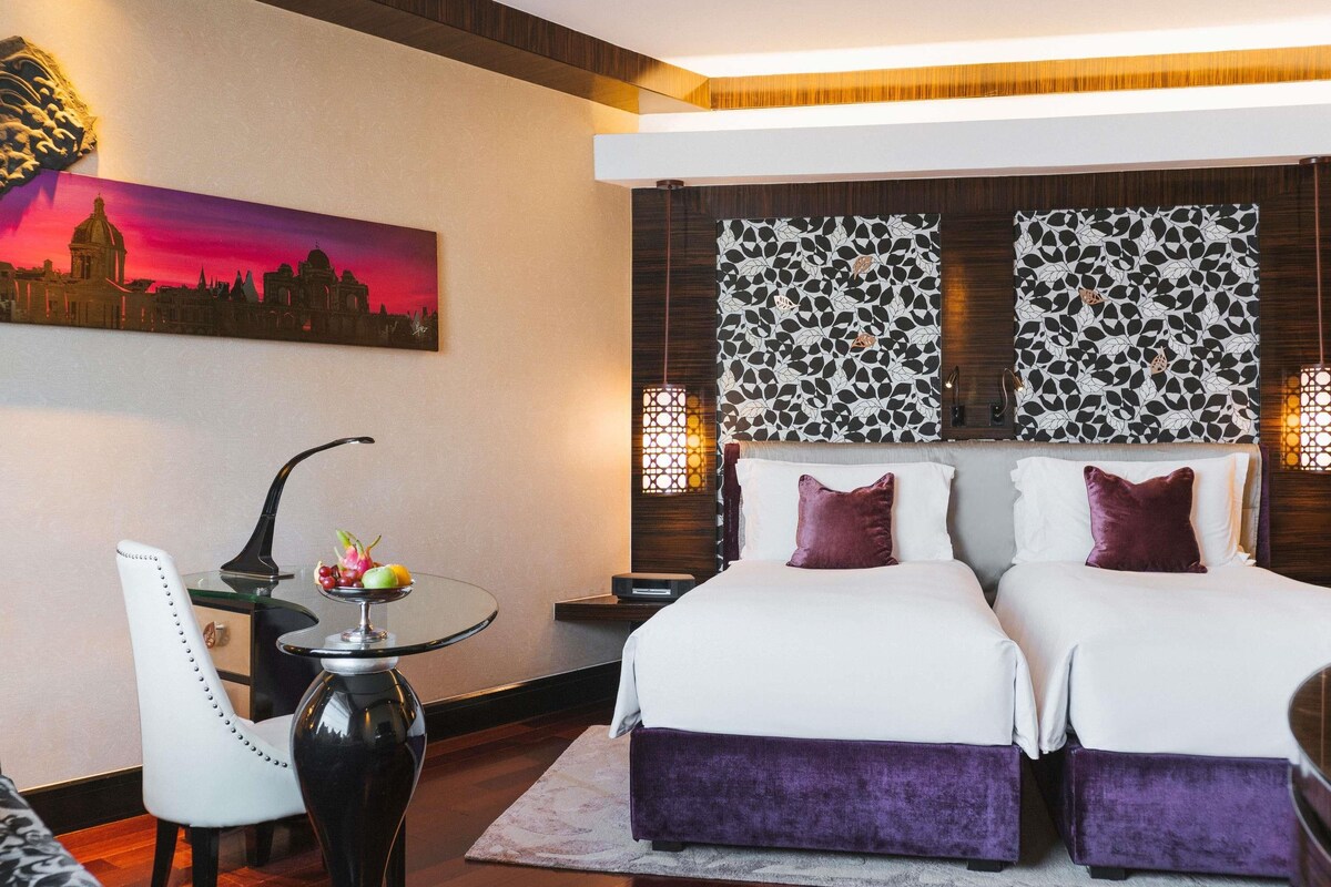 Quarto de hotel com duas camas, uma escrivaninha com um vaso, uma cadeira e um quadro com uma vista urbana na parede.