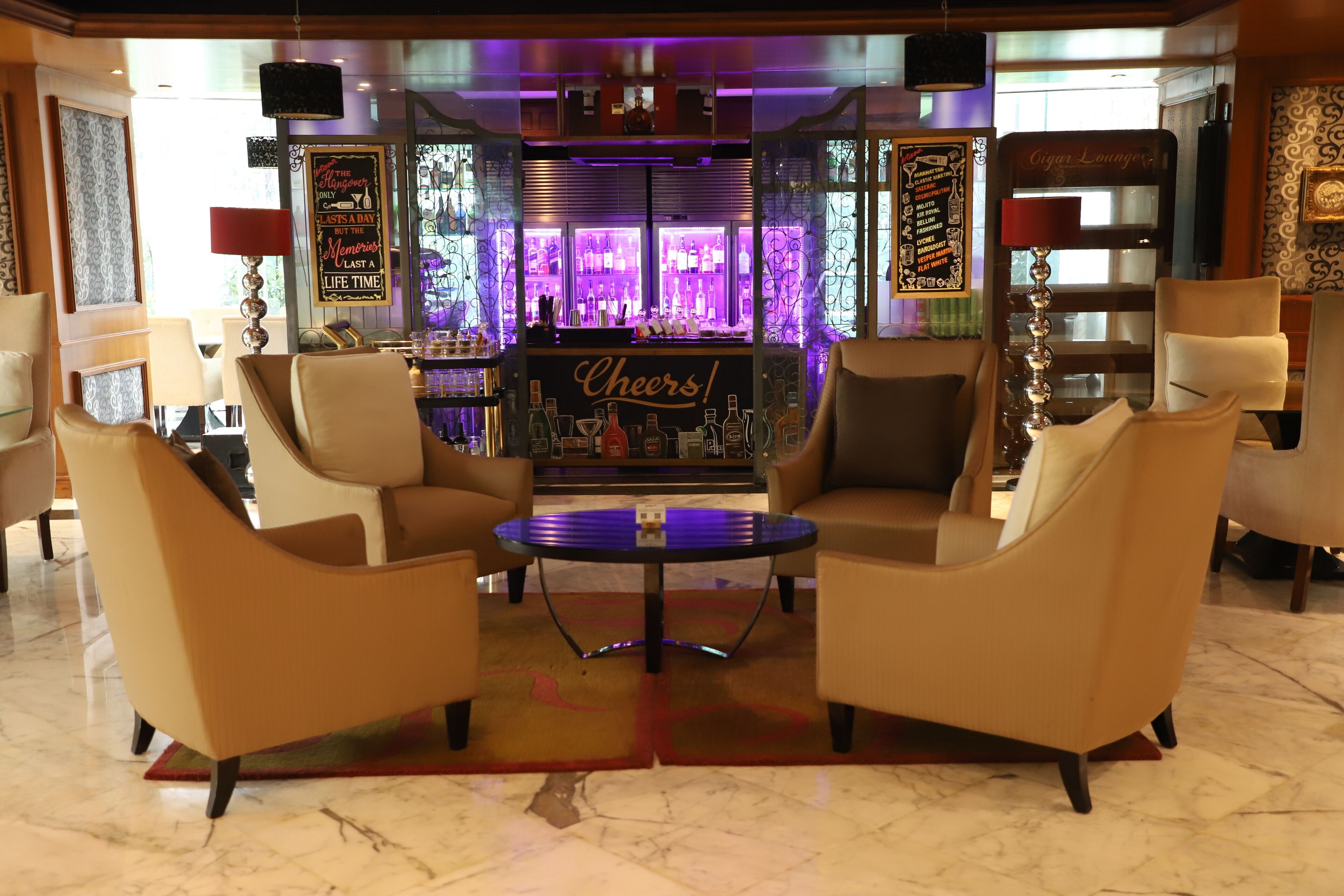Uma área de bar com um letreiro que diz 'Cheers!', duas poltronas bege, piso de mármore e uma divisória de vidro com prateleiras iluminadas.