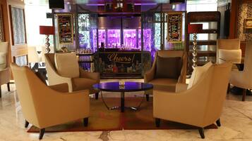 Uma área de bar com um letreiro que diz 'Cheers!', duas poltronas bege, piso de mármore e uma divisória de vidro com prateleiras iluminadas.
