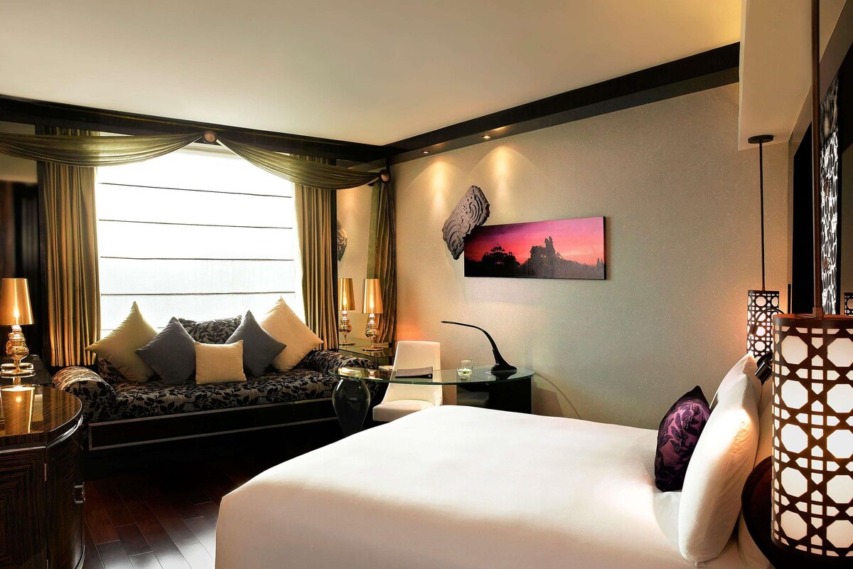 Quarto de hotel com uma cama grande, um sofá, uma escrivaninha com abajur e um quadro na parede.